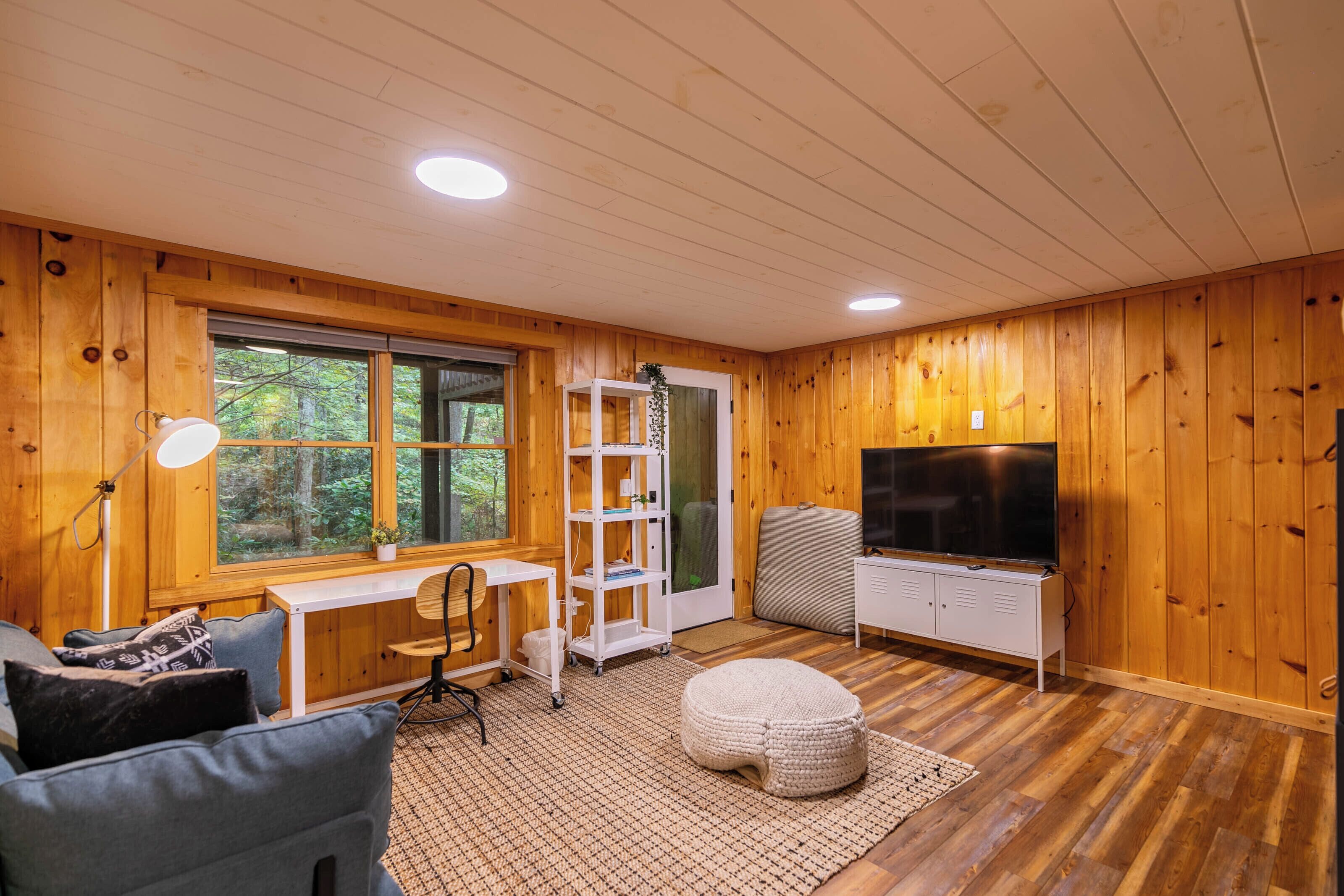 Quarto com painéis de madeira, uma escrivaninha, cadeira, apoio para os pés, televisão e janela com vista para uma floresta.