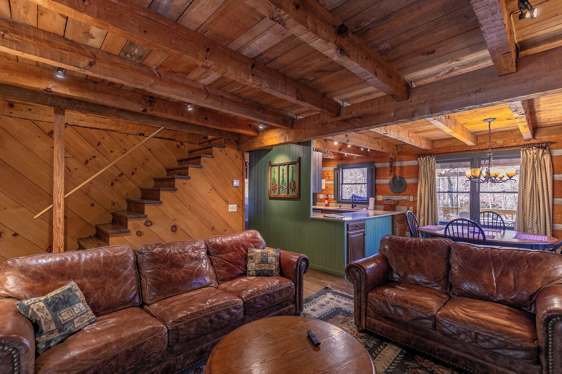 Interior acolhedor de uma cabana, com vigas de madeira, sofás de couro, sala de jantar e cozinha.
