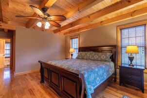 Quarto com teto de madeira, cama com colcha estampada, duas luminárias de cabeceira e janela com persianas.