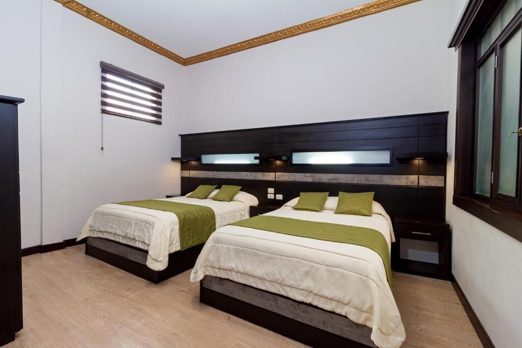 Quarto de hotel com duas camas, cabeceira escura, janela com persianas e piso de madeira.