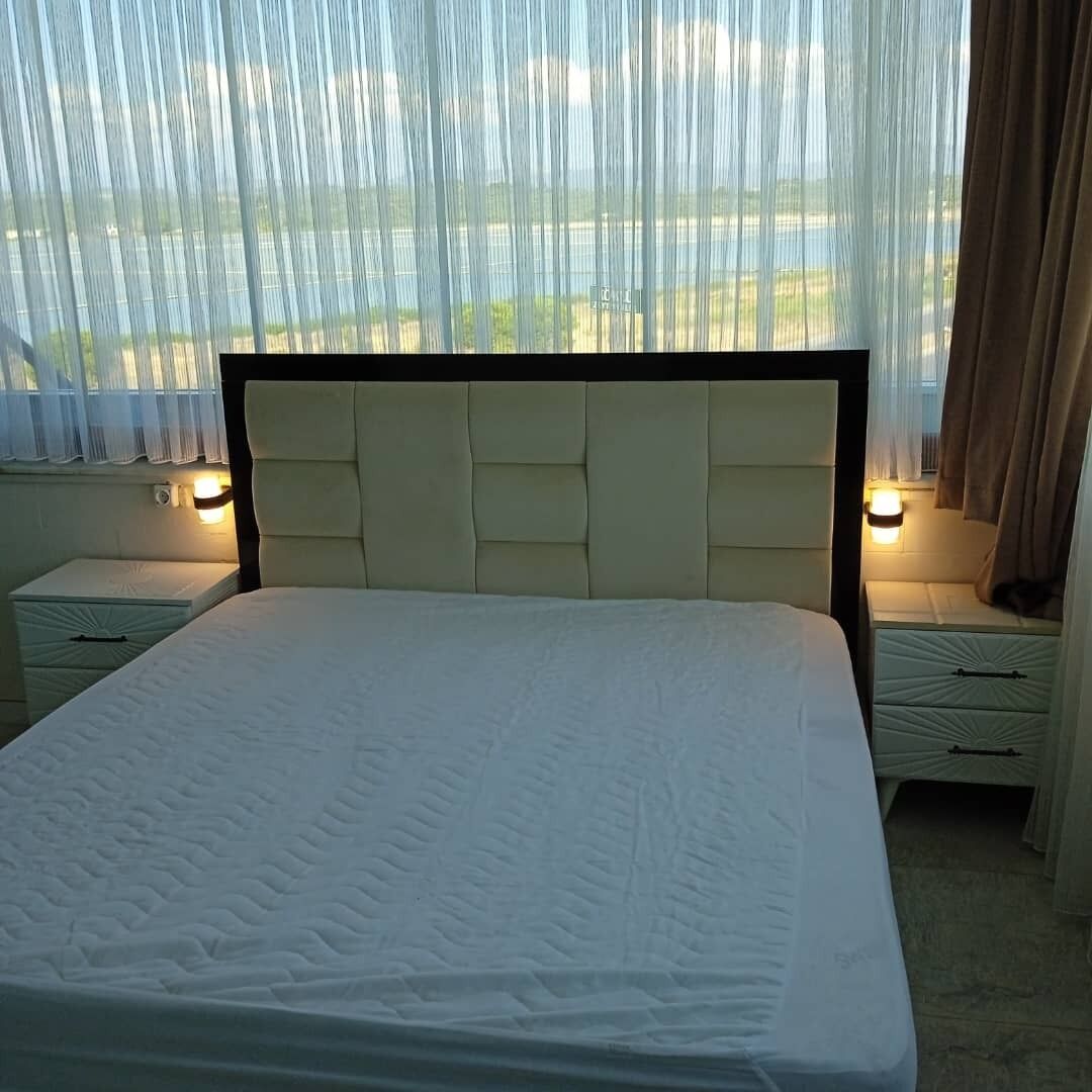 Uma cama com cabeceira, duas mesas de cabeceira com luminárias e vista para uma paisagem através de cortinas leves.