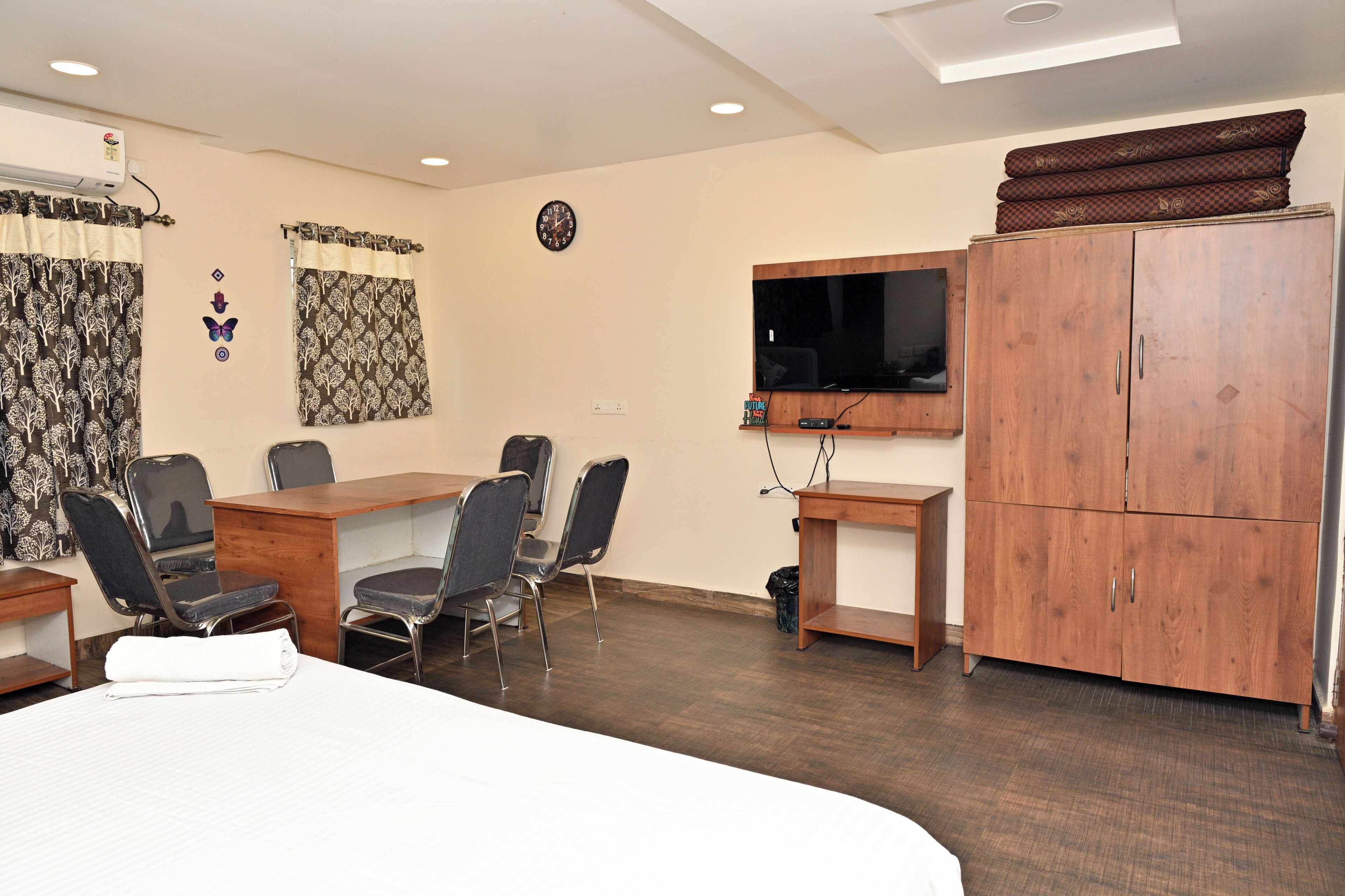 Quarto de hotel com cama, escrivaninha com cadeiras, televisão fixada na parede e guarda-roupa de madeira.