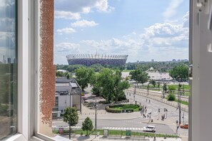 Visão de um estádio moderno com uma estrutura de cobertura única, cercado por um horizonte urbano com ruas, áreas verdes e céu claro.