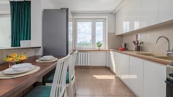 Uma cozinha moderna com armários brancos, uma mesa de jantar posta com pratos e uma fruteira, uma janela com vista para a cidade e um radiador sob a janela.