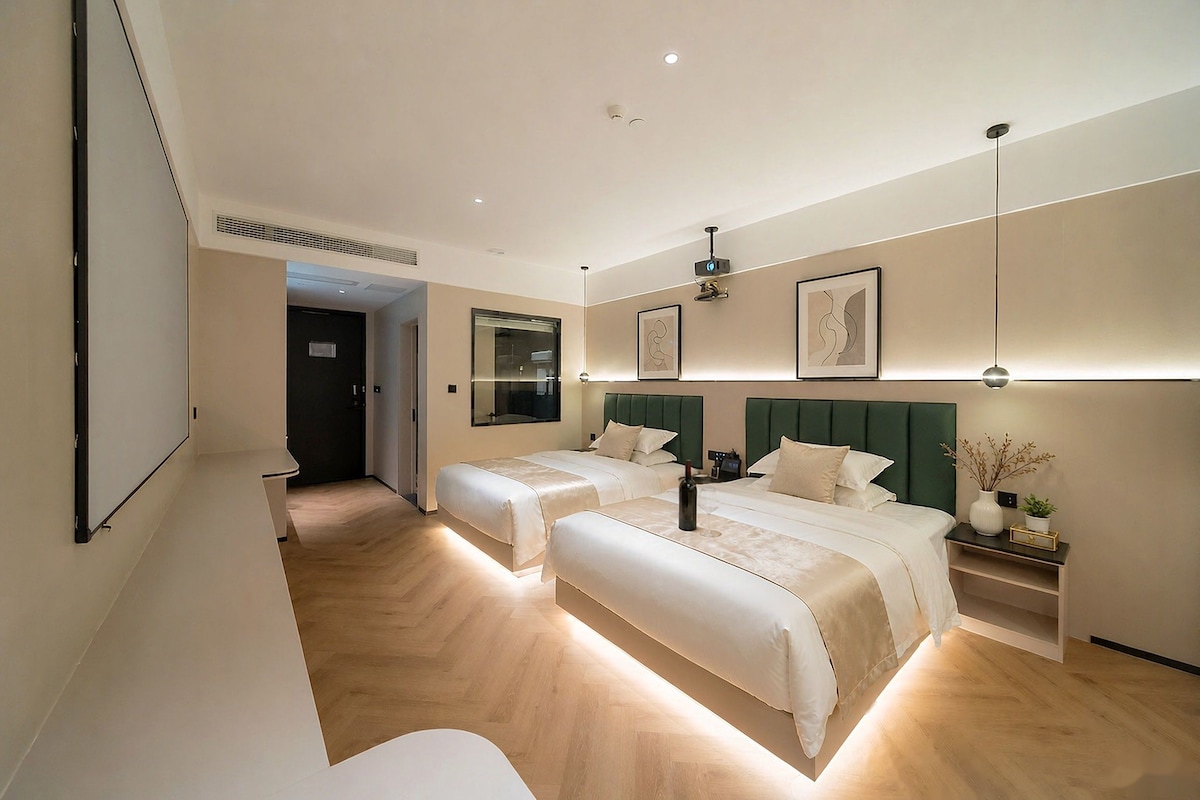 Quarto de hotel com duas camas, uma mesa de cabeceira com abajur, TV fixada na parede e uma porta que dá para outro cômodo.