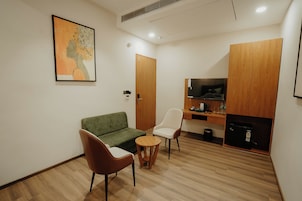 Um espaço de estar compacto com um sofá verde, duas poltronas, uma mesinha redonda, um armário de madeira e uma televisão.
