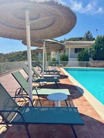 Uma área à beira da piscina com espreguiçadeiras e guarda-sóis, uma piscina e um edifício ao fundo.