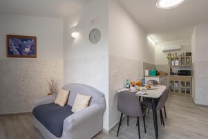 Uma sala de estar com sofá, uma mesa de jantar com cadeiras, uma cozinha e um relógio na parede.