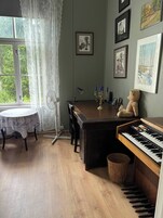 Um quarto com um piano, uma mesa de jantar com cadeiras, um ventilador e um urso de pelúcia.