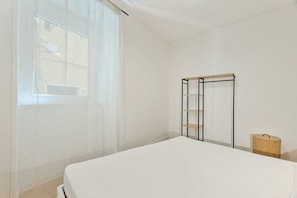 Quarto com cama, janela com cortinas leves, guarda-roupa de metal e cesto de vime.