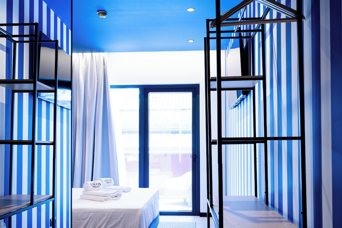 Quarto de hotel moderno com beliches, uma janela grande e vista para a cidade.