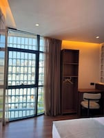 Quarto de hotel com cama, escrivaninha, cadeira, guarda-roupa e uma grande janela com vista para edifícios.