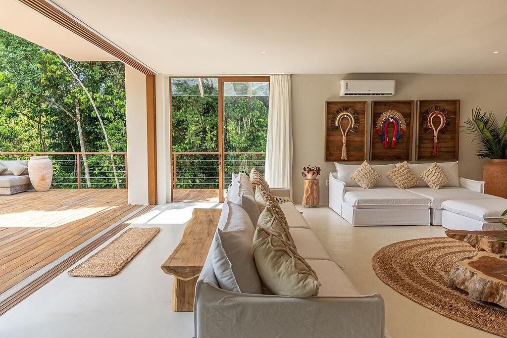Uma sala de estar moderna com um sofá grande, uma mesa de centro de madeira e vista para o verde exuberante através de grandes portas deslizantes.