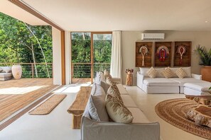 Uma sala de estar moderna com um sofá grande, uma mesa de centro de madeira e vista para o verde exuberante através de grandes portas deslizantes.