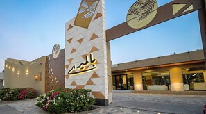 Entrada de hotel com fachada geométrica, sinalização iluminada e área de acesso ajardinada.