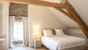 Quarto com teto inclinado, uma cama, mesa de cabeceira e janela com cortinas.