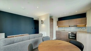 Uma sala de estar moderna com um sofá cinzento, uma mesa de jantar de madeira e uma cozinha com armários de madeira e forno embutido.