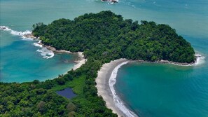 Vista aérea de uma ilha tropical com uma praia, rodeada por águas azul-claras e vegetação verde exuberante.
