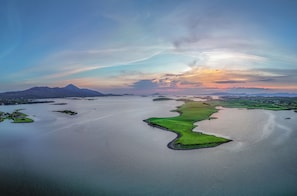 Paisagem costeira com uma grande extensão de água, áreas verdes e montanhas ao longe sob um céu claro.