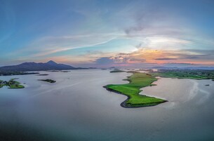 Paisagem costeira com uma grande extensão de água, áreas verdes e montanhas ao longe sob um céu claro.