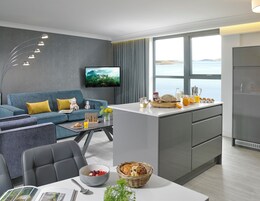 Uma sala de estar moderna com sofá, mesa de jantar com itens de café da manhã, cozinha com geladeira e uma grande janela com vista.