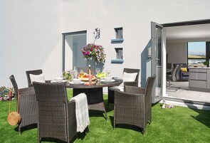 Uma área de refeições ao ar livre com móveis de vime, uma mesa posta com pratos, copos e um cesto de flores pendurado.