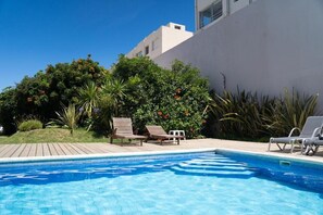 Piscina com espreguiçadeiras, cercada por vegetação e um edifício moderno.
