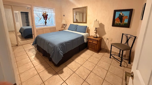 Quarto com cama, mesa de cabeceira, cadeira, ventilador de teto, janela com persianas, espelho e um quadro na parede.