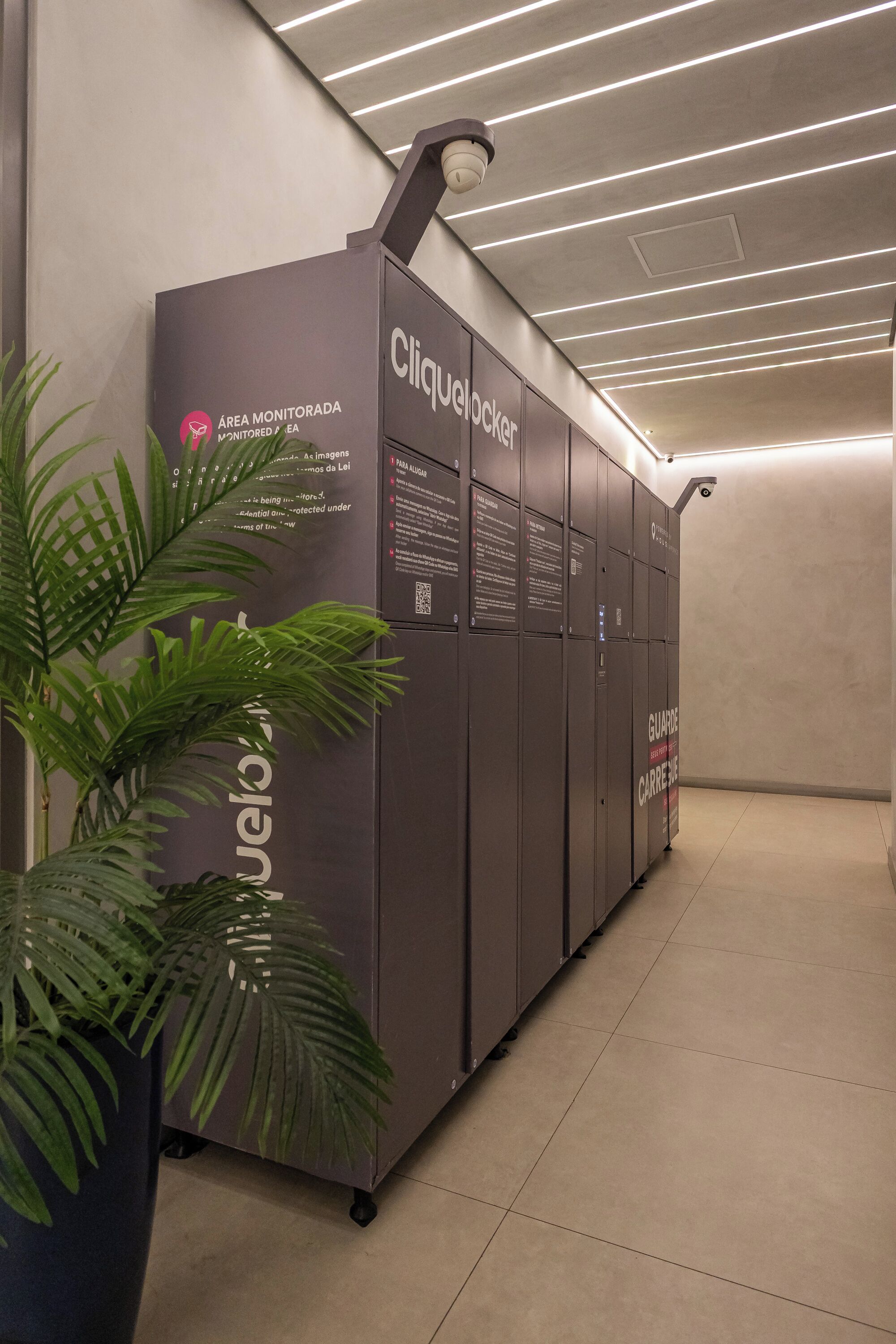 Um elevador moderno com a marca 'Cliclocker', um vaso de planta e sinalização informativa.