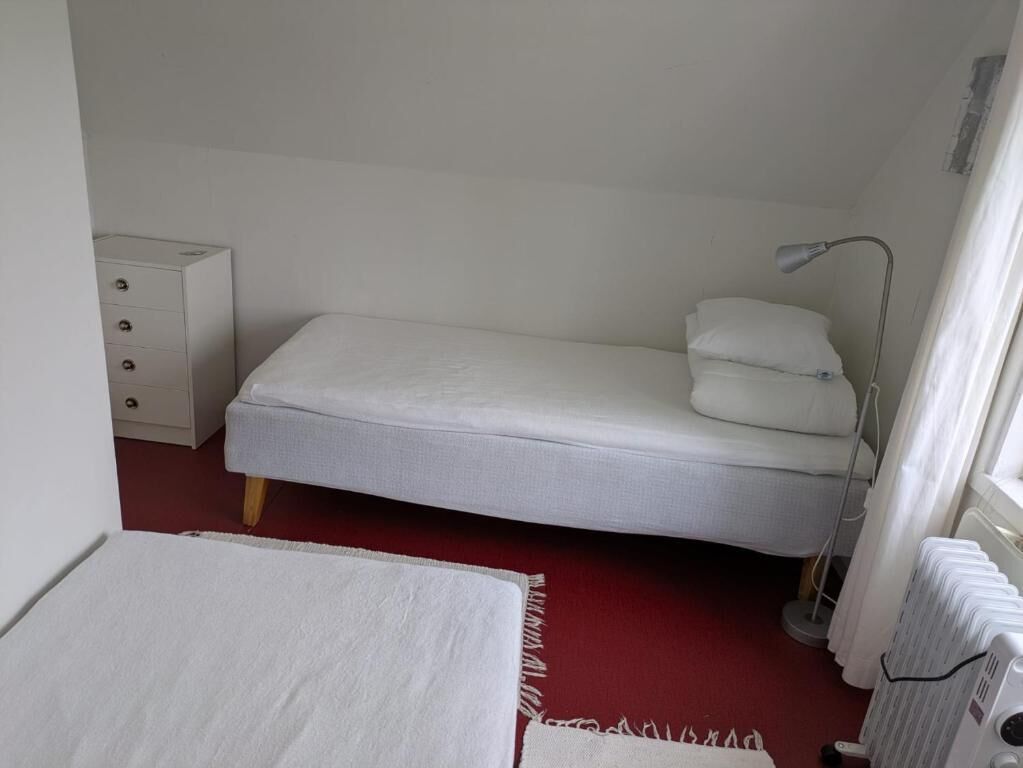Quarto pequeno com uma cama de solteiro, colchão branco, luminária de cabeceira, radiador branco e cômoda pequena e branca.