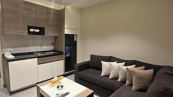 Uma sala de estar moderna com um sofá cinza escuro, uma mesa de centro branca e uma área de cozinha com armários de madeira e micro-ondas embutido.