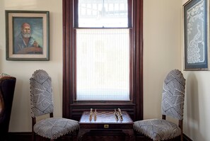Um quarto com um tabuleiro de xadrez, duas poltronas estampadas, um retrato emoldurado e uma janela com persianas.