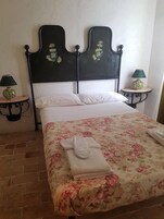 Uma cama com colcha florida, cabeceira escura com detalhes florais, duas mesas de cabeceira com abajures e um chão de tijolos.