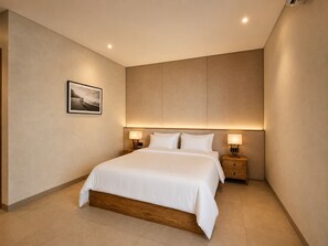 Quarto de hotel moderno com uma cama grande, duas mesas de cabeceira com abajures, uma moldura na parede e uma paleta de cores neutras.