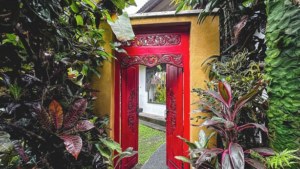 Uma porta vermelha que dá para um edifício com parede amarela, rodeado por vegetação exuberante.