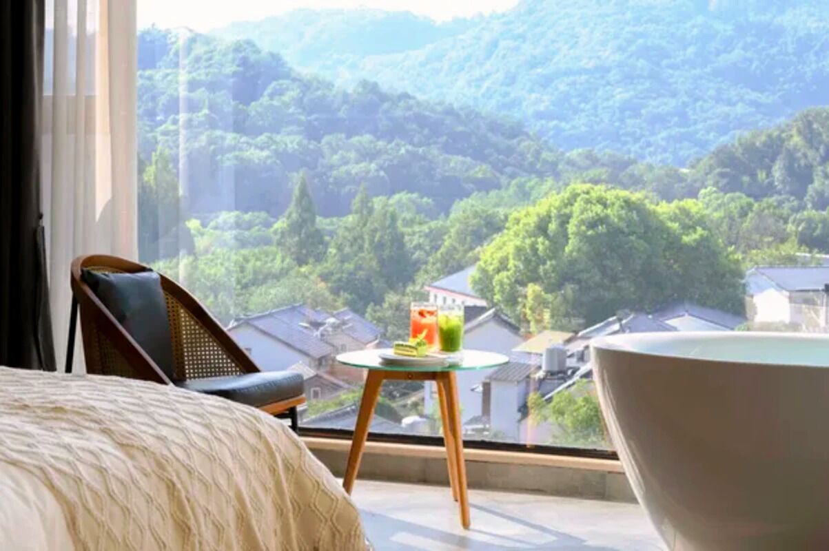 Quarto com cama, cadeira, mesa com bebida e vista para uma área arborizada.