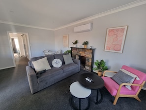 Uma sala de estar com um sofá cinza, uma mesa redonda pequena, uma poltrona rosa, uma lareira e um ar-condicionado fixado na parede.