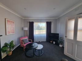 Quarto com televisão, uma cadeira rosa, mesa redonda, planta em vaso e uma janela grande com cortinas.