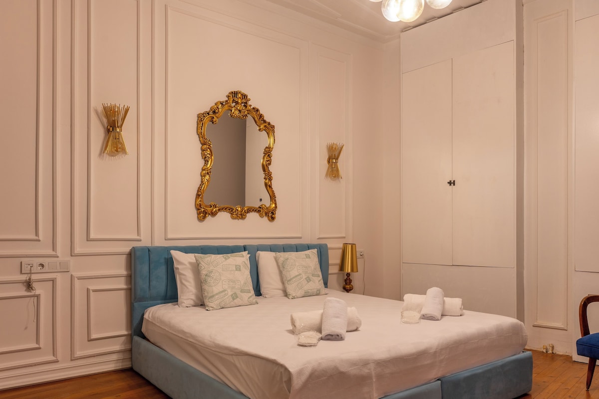Quarto de hotel com uma cama grande, um espelho com moldura dourada e cabeceira azul.