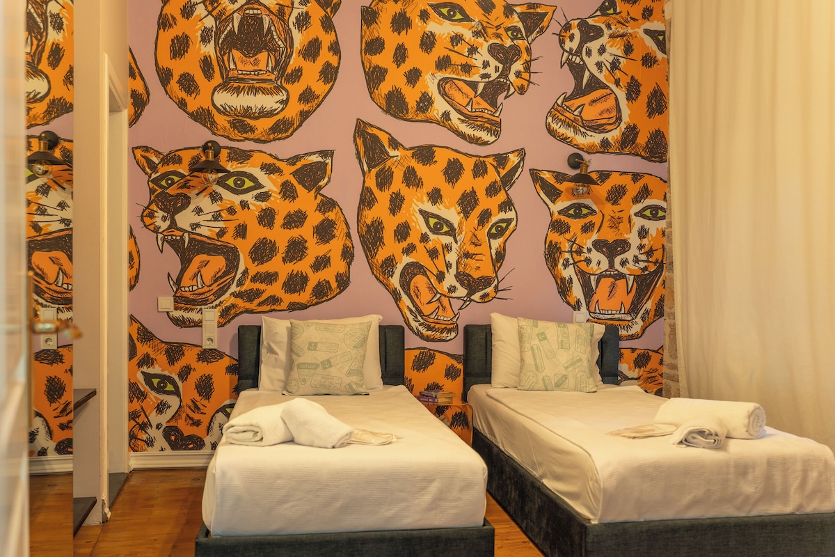 Quarto de hotel com duas camas, parede grande com estampa de leopardo e piso de madeira.