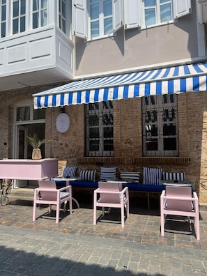 Um edifício com fachada de pedra, toldo riscado em azul e branco, e área de estar com almofadas riscadas e cadeiras cor-de-rosa.