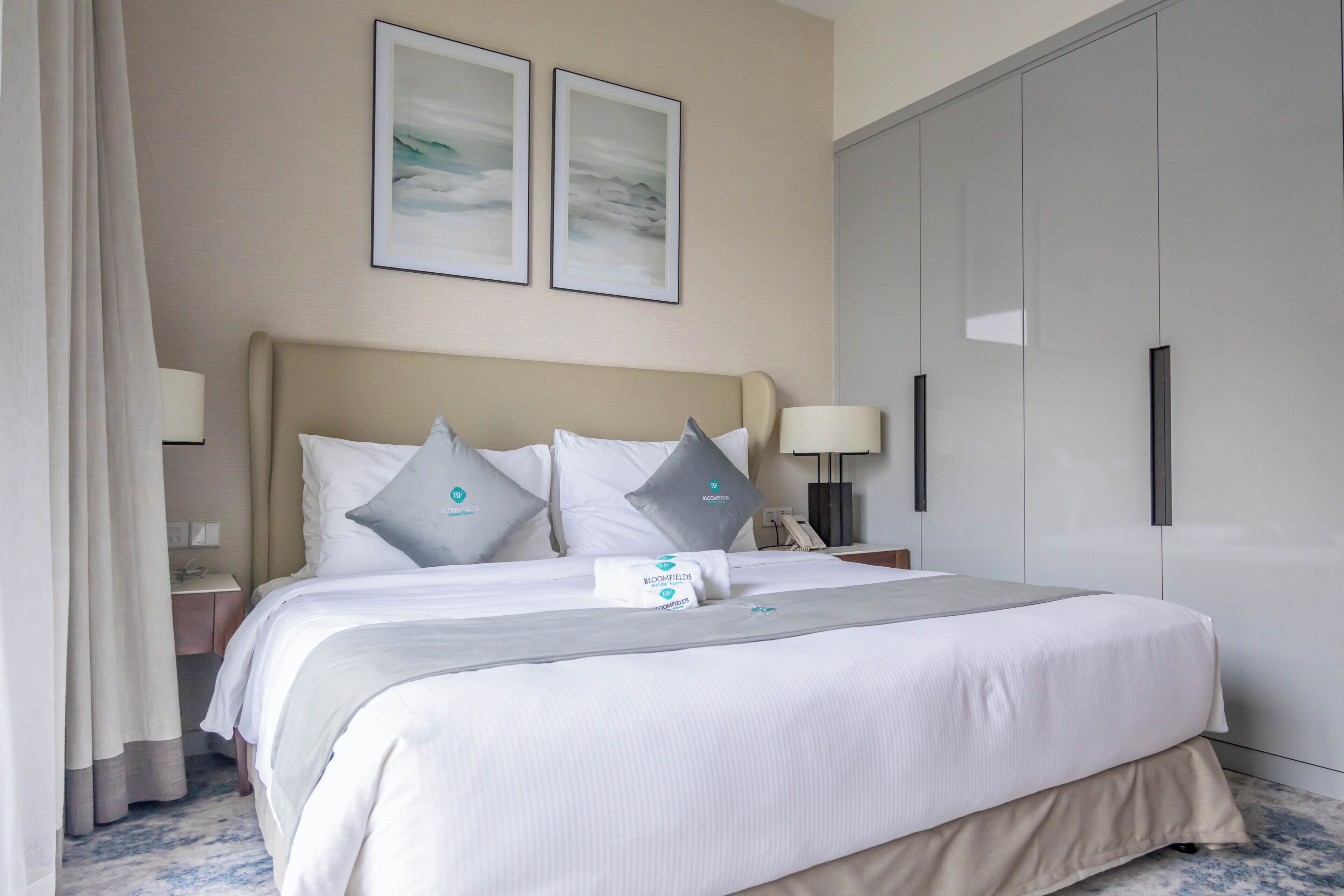 Quarto de hotel com cama, mesa de cabeceira, abajur, guarda-roupa e duas imagens emolduradas na parede.