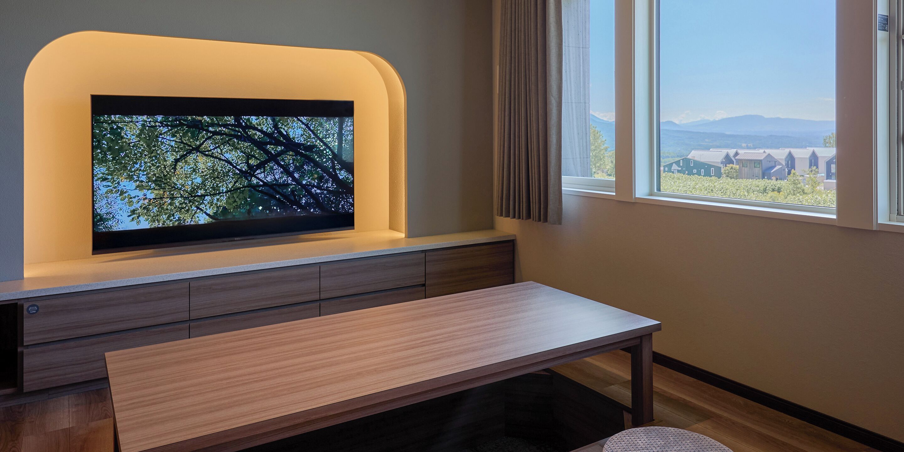 Quarto moderno com televisão de tela plana na parede, um banco de madeira e uma janela com vista para casas e montanhas.