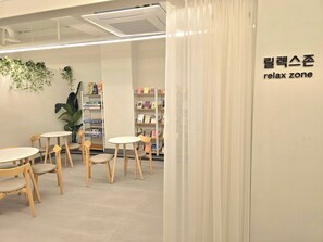 Um espaço interno tranquilo com mesas, cadeiras, uma estante de livros e uma placa indicando uma 'zona de relaxamento'.