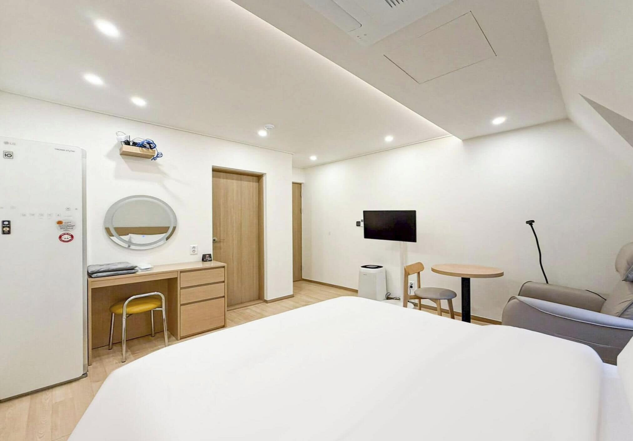 Quarto de hotel moderno com cama, escrivaninha com cadeira, espelho redondo, televisão fixada na parede e pequena mesa redonda com cadeira.