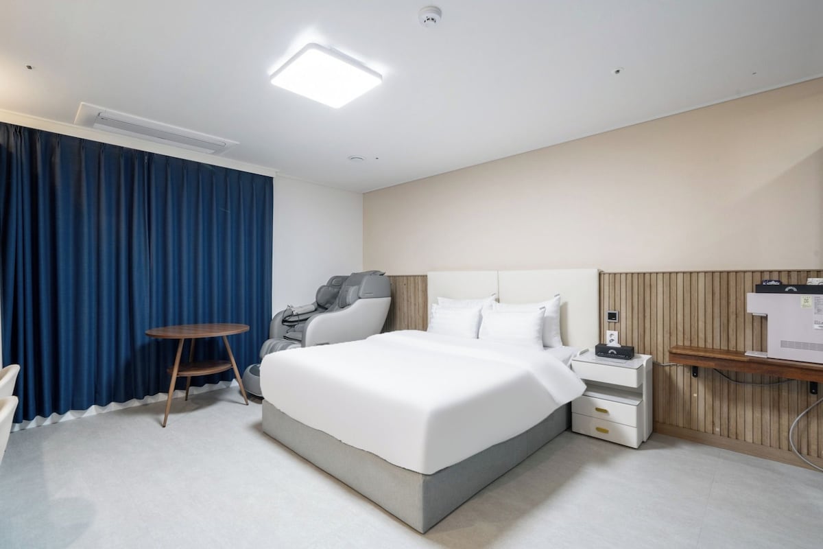 Quarto de hotel com cama, cadeira de massagem, mesa pequena, escrivaninha e aquecedor fixado na parede.