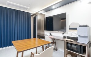Quarto de hotel com mesa de madeira, cadeiras brancas, televisão de tela plana, minibar e máquina de café.