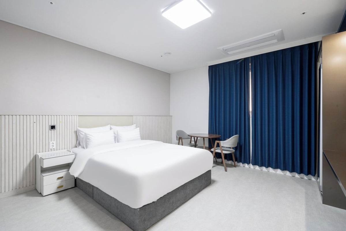 Quarto de hotel moderno com cama grande, mesas de cabeceira, escrivaninha com cadeiras e janela com cortinas azuis.