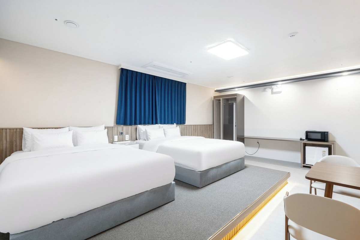 Quarto de hotel com duas camas, uma escrivaninha, uma cadeira, micro-ondas e uma porta.