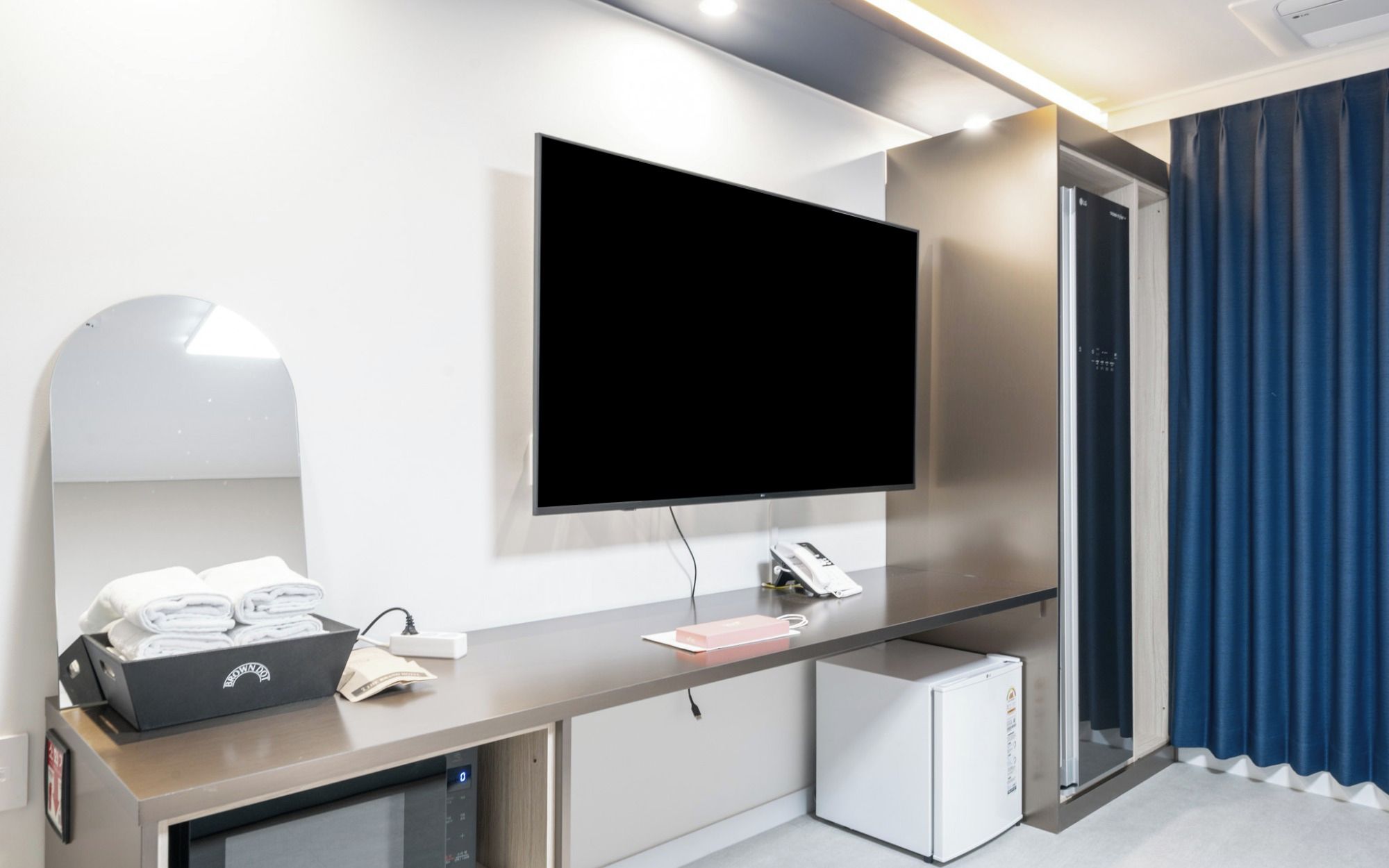 Quarto de hotel com televisão de tela plana na parede, uma pequena escrivaninha com telefone, um mini-refrigerador e uma caixa de artigos de higiene pessoal.